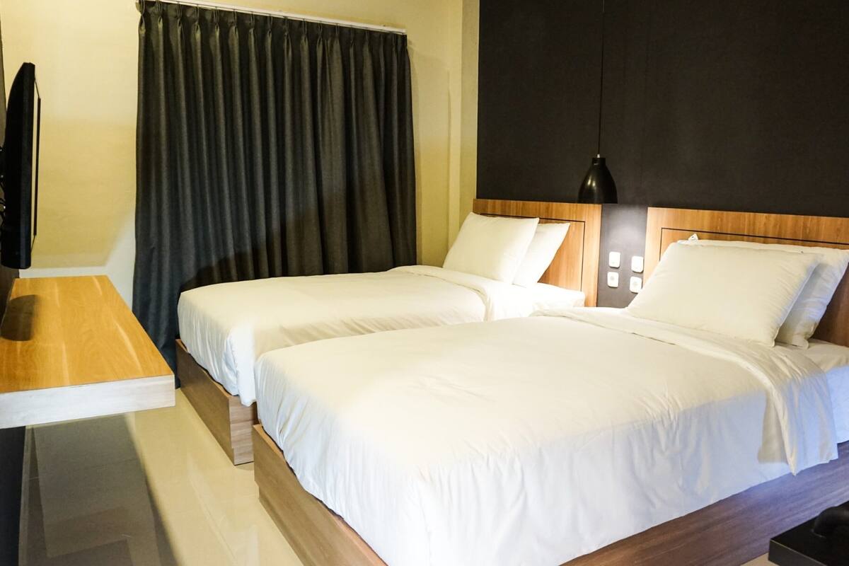 Quarto de hotel com duas camas, uma mesa de cabeceira de madeira, uma televisão e uma cortina escura.