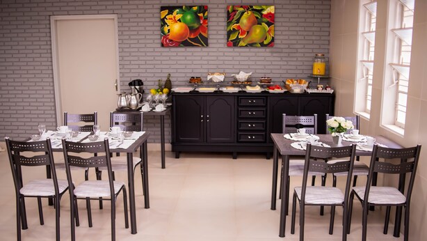 Uma área de refeições com parede de tijolos, duas pinturas de frutas, um armário preto com gavetas e uma janela com persianas.