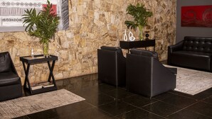 Um lobby com paredes de pedra, sofás de couro preto e duas mesinhas laterais com plantas.
