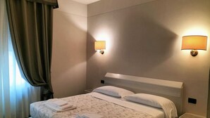 Quarto de hotel com uma cama, duas luminárias na parede, uma janela com cortinas e uma mesa de cabeceira.