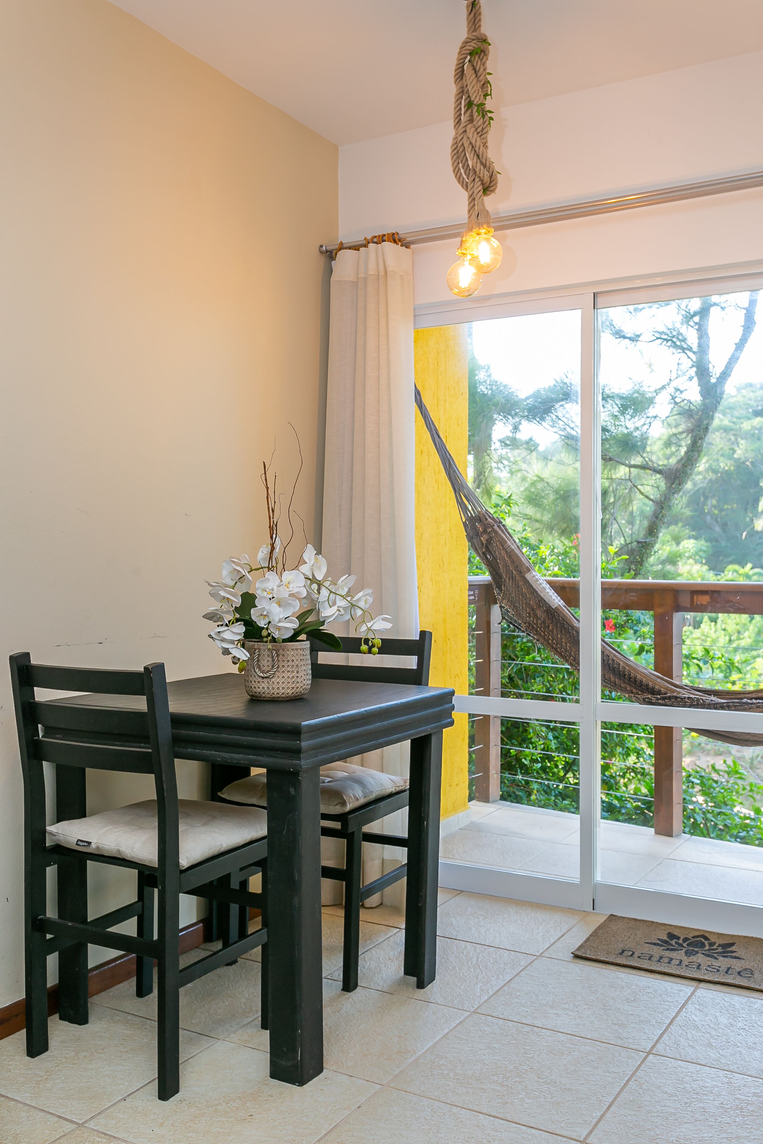 Uma área de refeições com mesa e cadeiras, uma rede pendurada e vista para o verde através de uma janela.