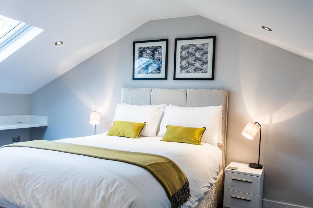 Quarto moderno com teto inclinado, cama com roupa de cama branca e duas almofadas, mesa de cabeceira com abajur e duas obras de arte emolduradas na parede.