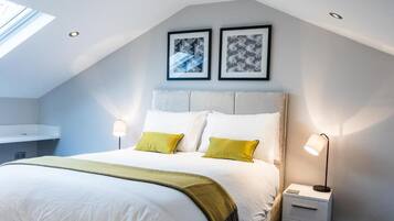 Quarto moderno com teto inclinado, cama com roupa de cama branca e duas almofadas, mesa de cabeceira com abajur e duas obras de arte emolduradas na parede.