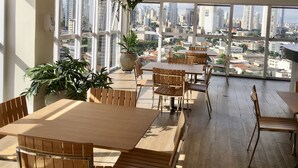 Uma sala de reuniões ampla com grandes janelas que oferecem vista para a cidade, mesas e cadeiras de madeira e plantas em vasos.
