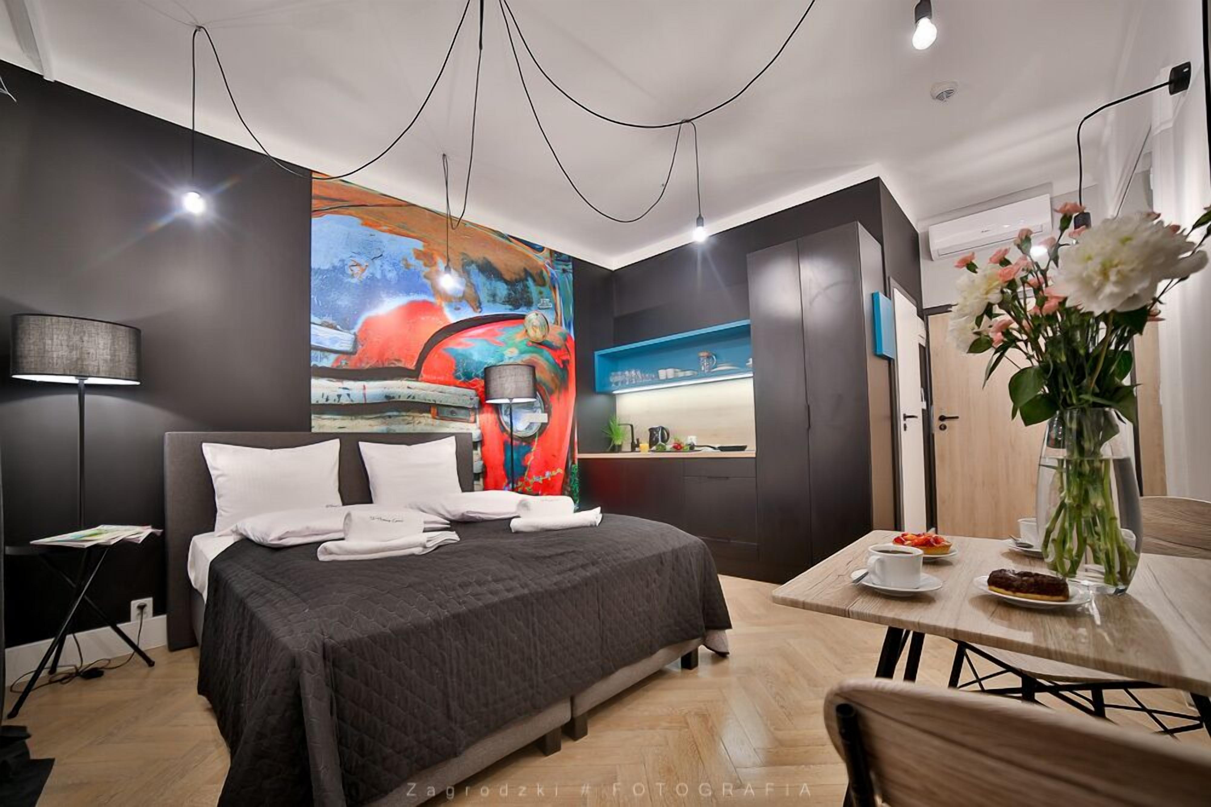 Quarto de hotel moderno com uma cama grande, uma escrivaninha com cadeira, uma mesa de jantar com um vaso de flores e um quadro abstrato colorido na parede.