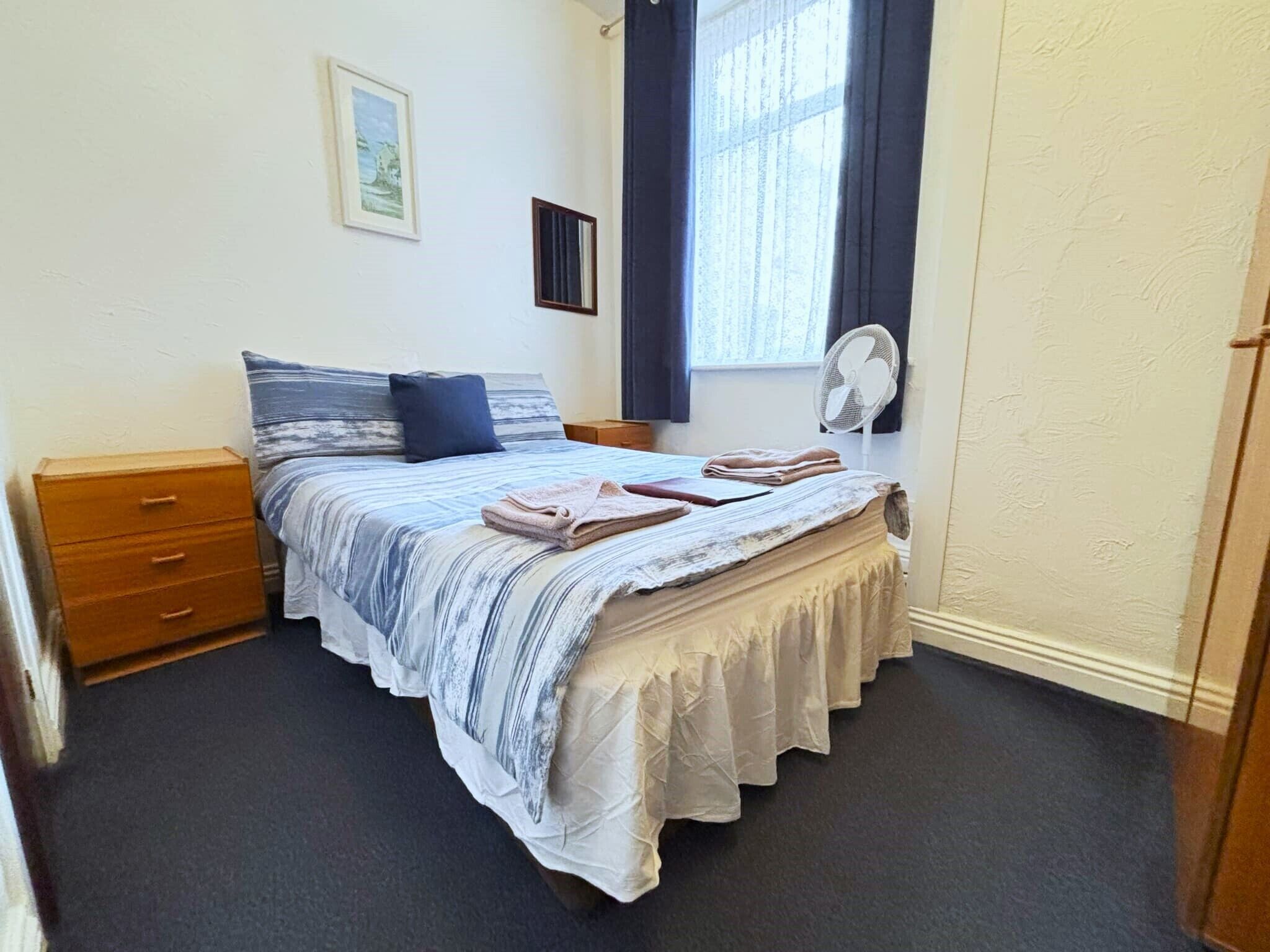 Quarto com cama, mesa de cabeceira, janela com cortinas, ventoinha fixada na parede e um pequeno porta-retrato.