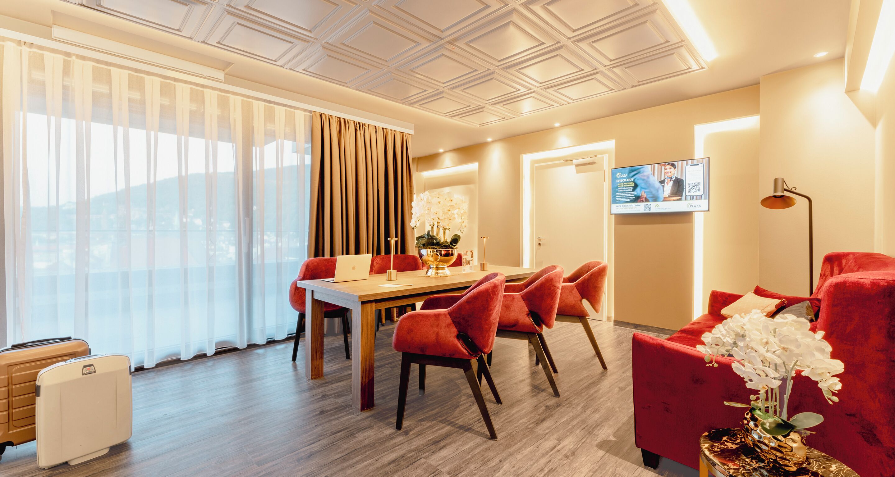 Quarto de hotel moderno com uma mesa de jantar de madeira, cadeiras estofadas vermelhas, um sofá vermelho, uma televisão na parede e uma grande janela com cortinas finas.