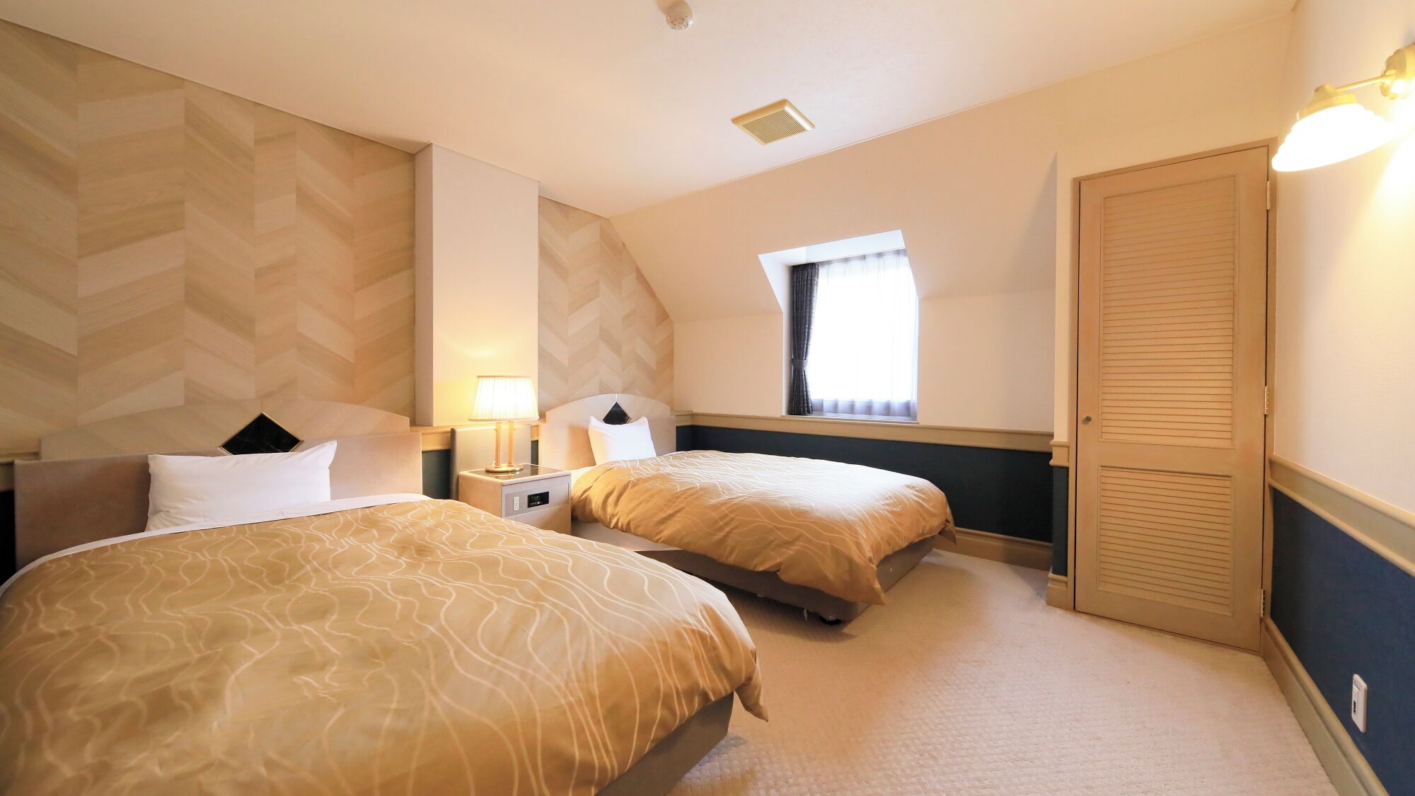 Quarto de hotel com duas camas, uma mesa de cabeceira, uma luminária, uma janela e uma porta.