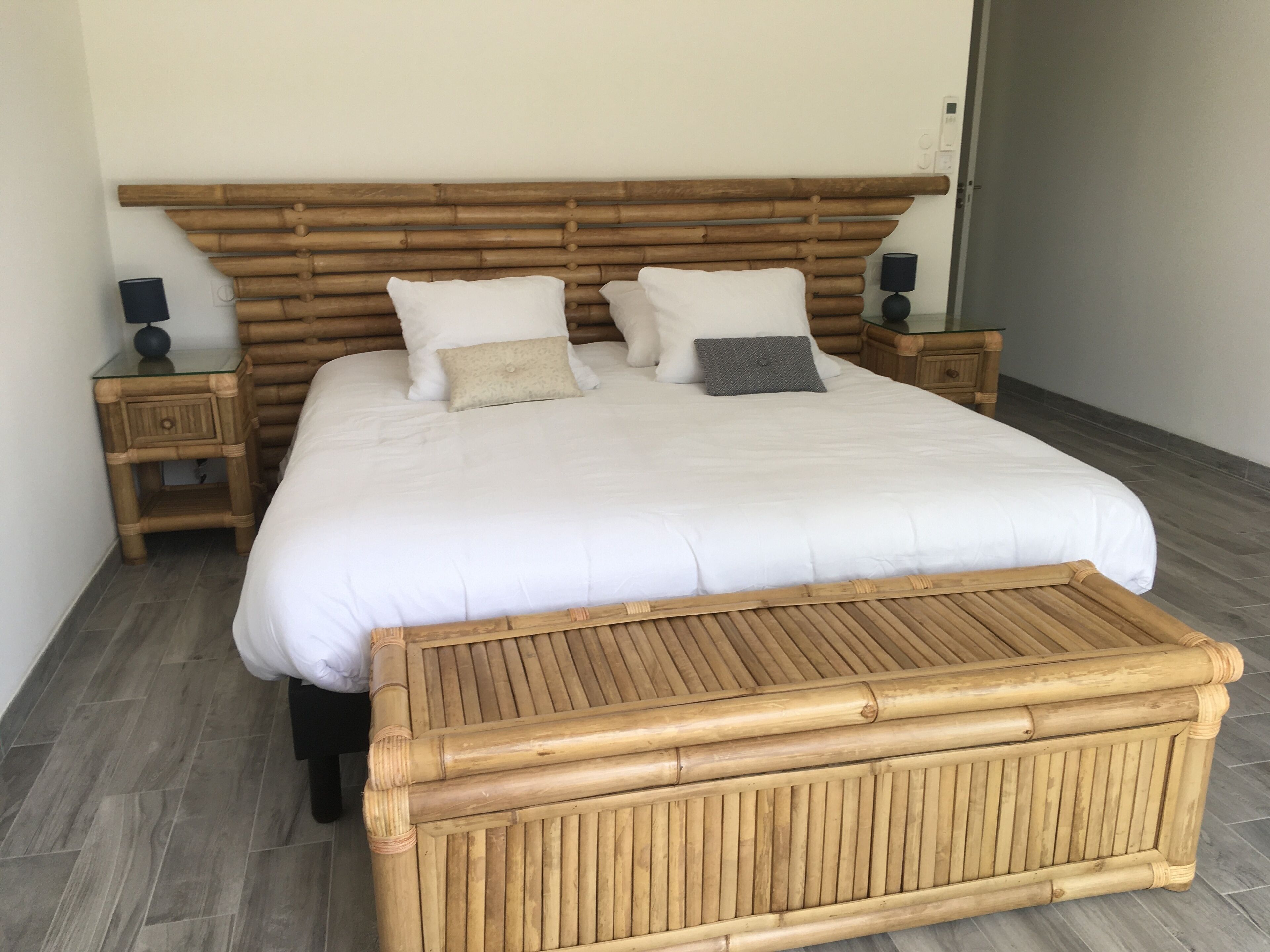 Uma cama bem arrumada com cabeceira de madeira, duas mesas de cabeceira com abajures e um banquinho de fibra natural no pé da cama.