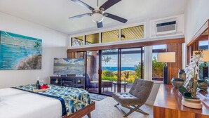 Quarto de hotel com cama, escrivaninha, cadeira, ventoinha no teto e uma grande porta de vidro deslizante que dá para uma varanda com vista para o oceano.