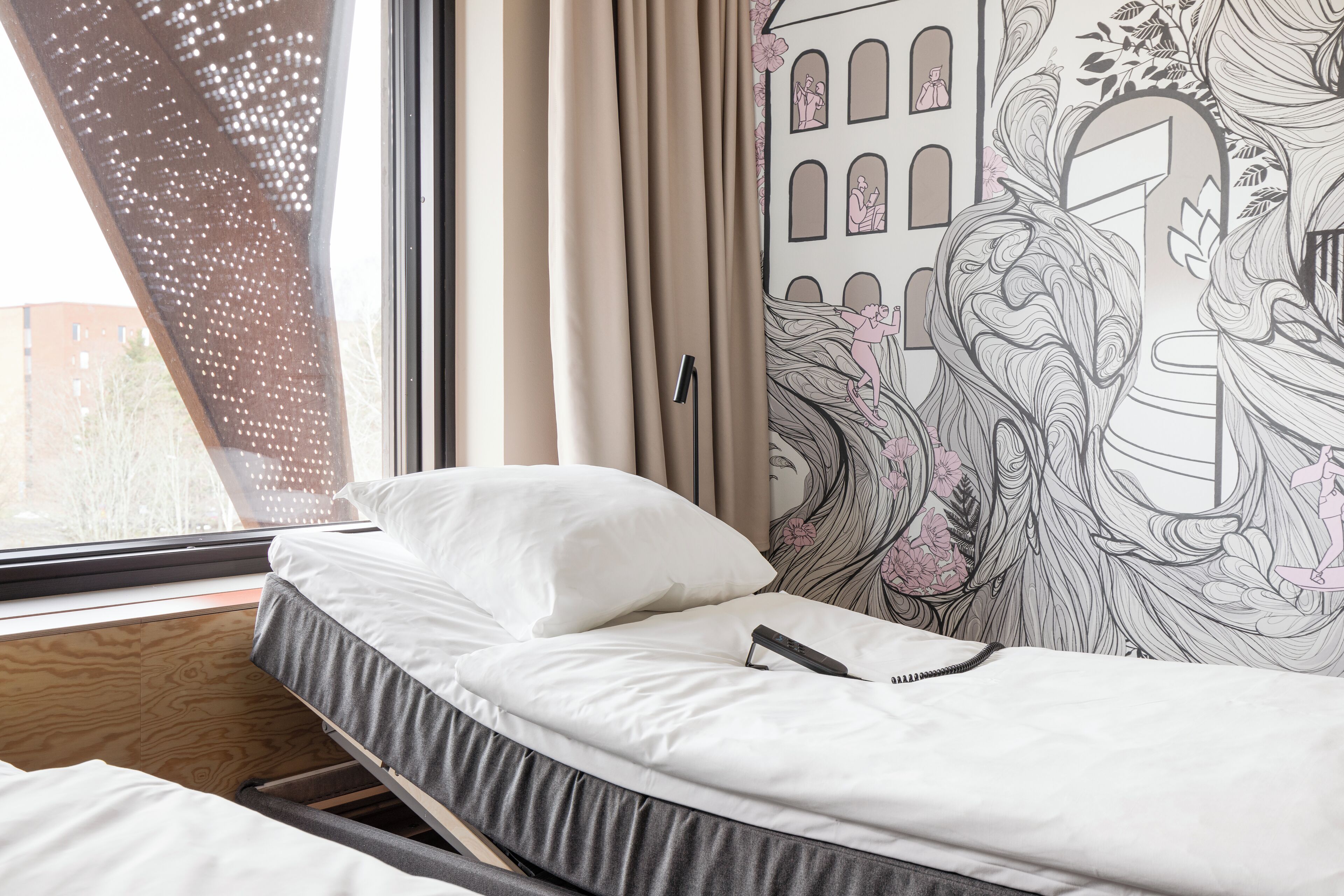 Uma cama com travesseiro branco e roupa de cama cinza, uma janela com cortina e uma parede com um mural detalhado.