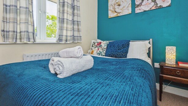 Quarto com cama, mesa de cabeceira, janela com cortinas e um painel decorativo com motivos florais.