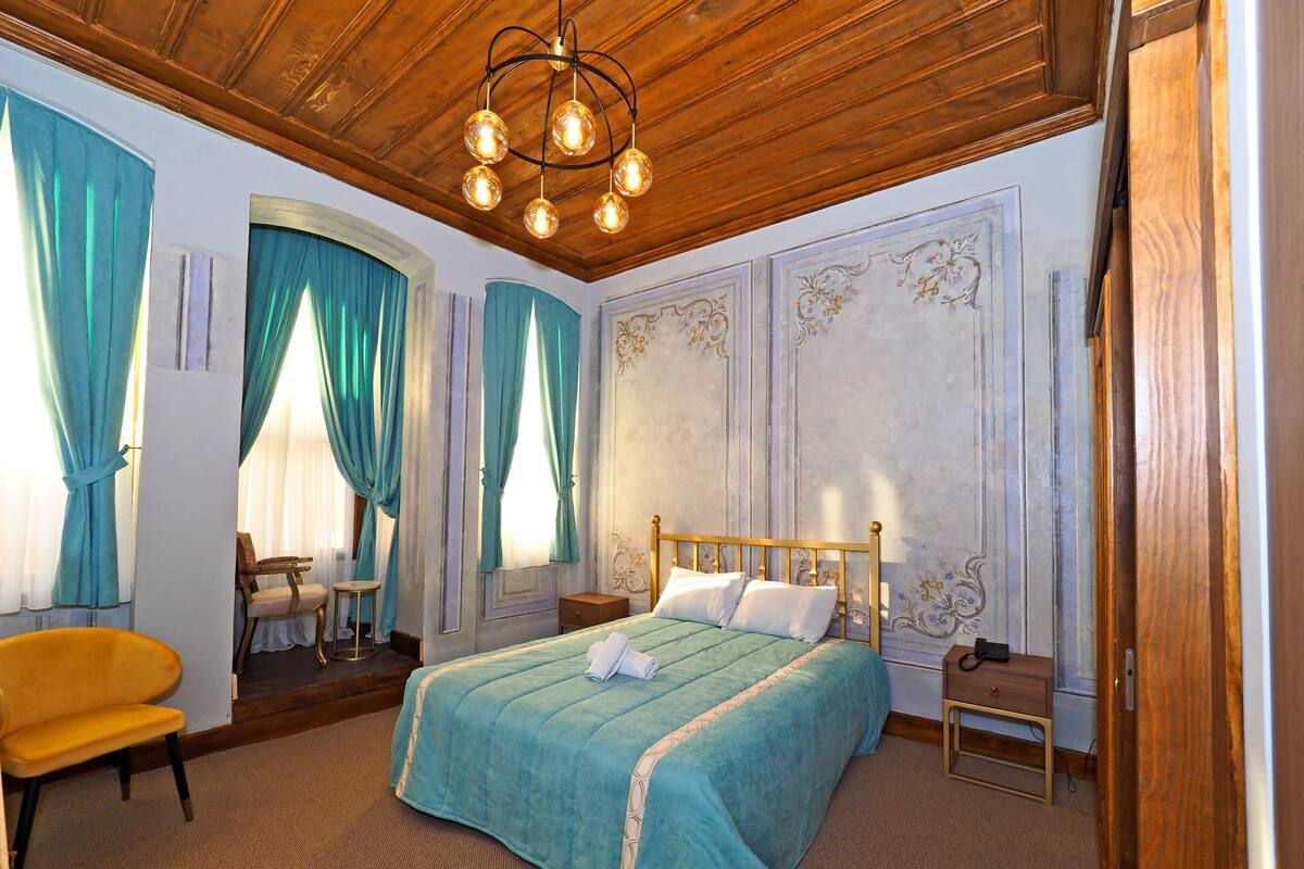 Quarto com teto de madeira, cama com colcha verde, duas mesas de cabeceira, uma cadeira e janela com cortinas.