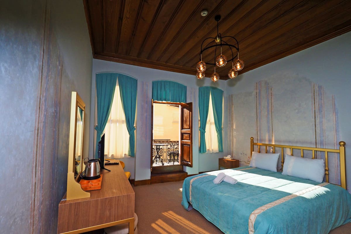 Quarto com teto de madeira, cama com colcha azul, escrivaninha com chaleira e janela com cortinas.