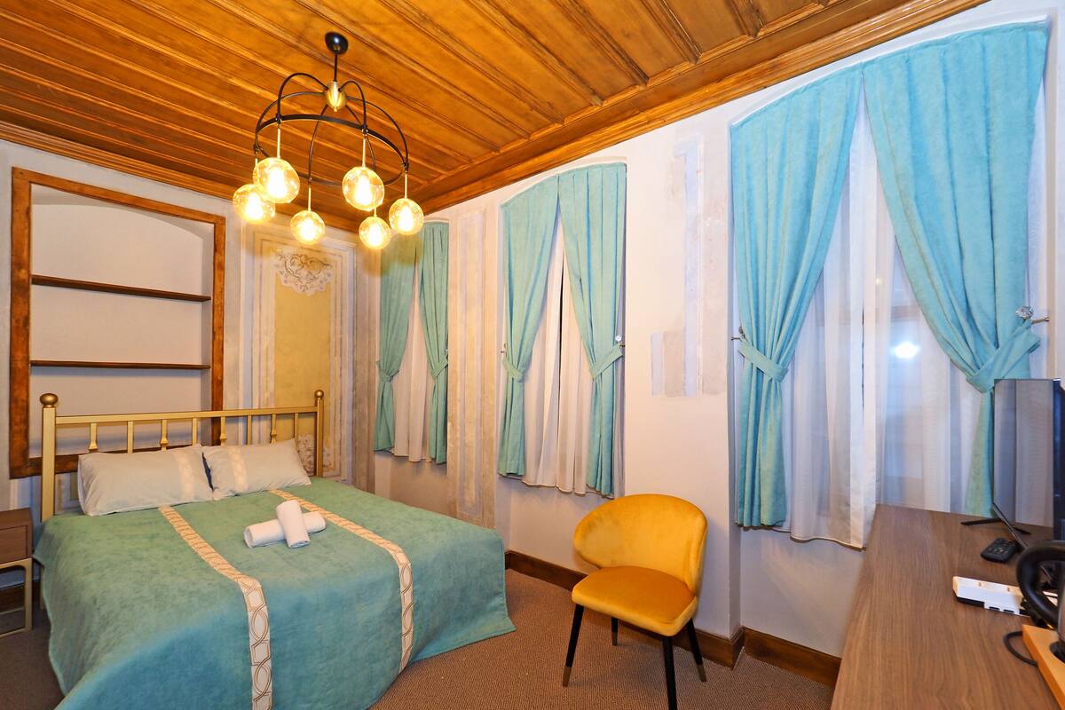 Quarto de hotel com teto de madeira, cama com colcha verde, poltrona amarela, escrivaninha com computador e cortinas nas janelas.