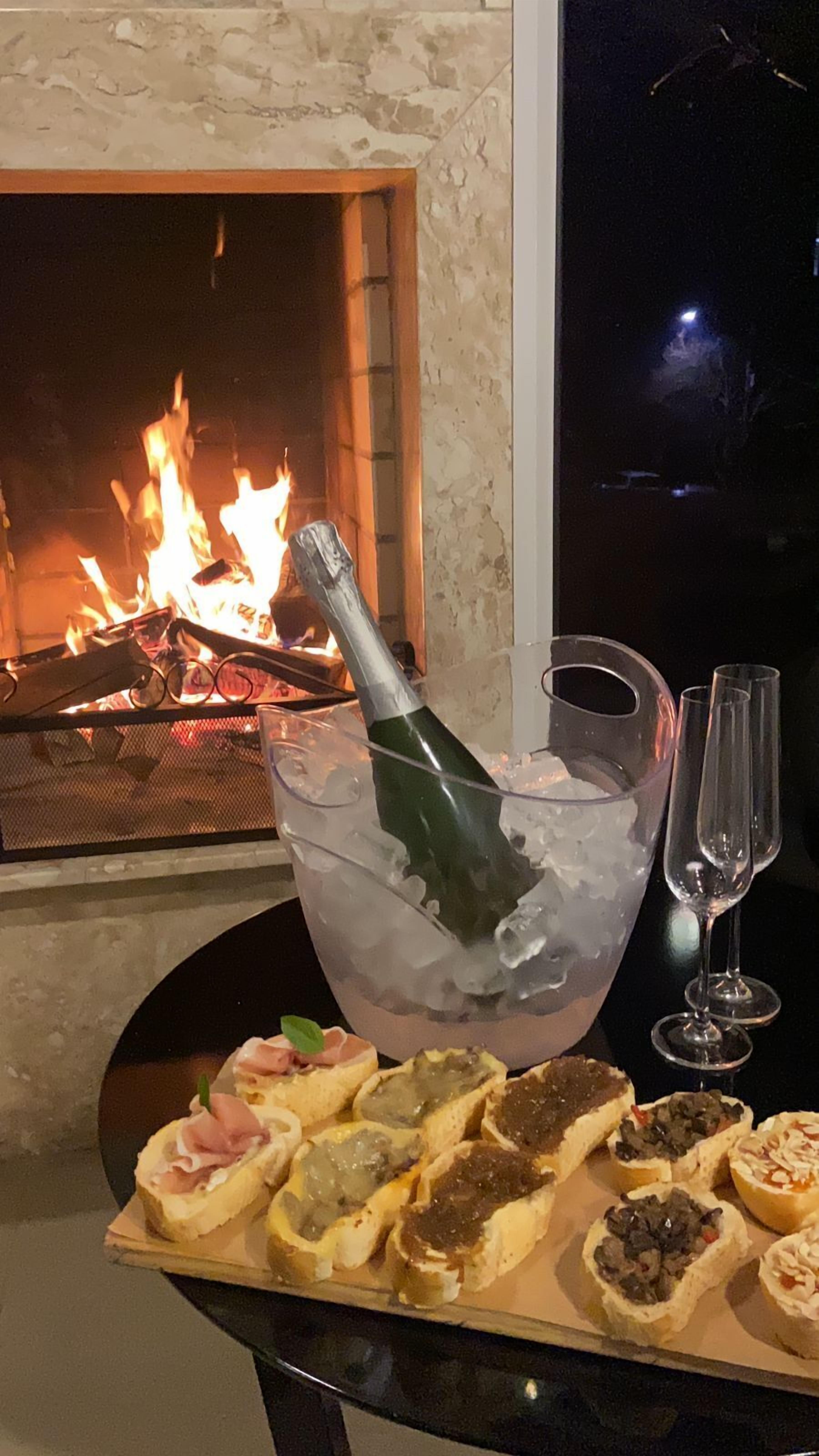Mesa com uma garrafa de champanhe em balde, rodeada por aperitivos, com uma lareira ao fundo.