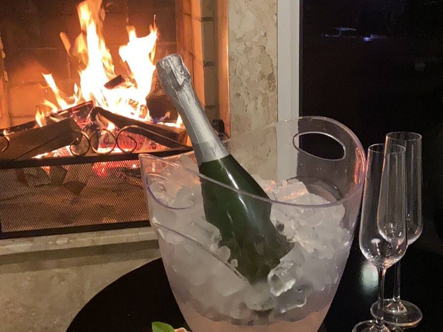 Mesa com uma garrafa de champanhe em balde, rodeada por aperitivos, com uma lareira ao fundo.