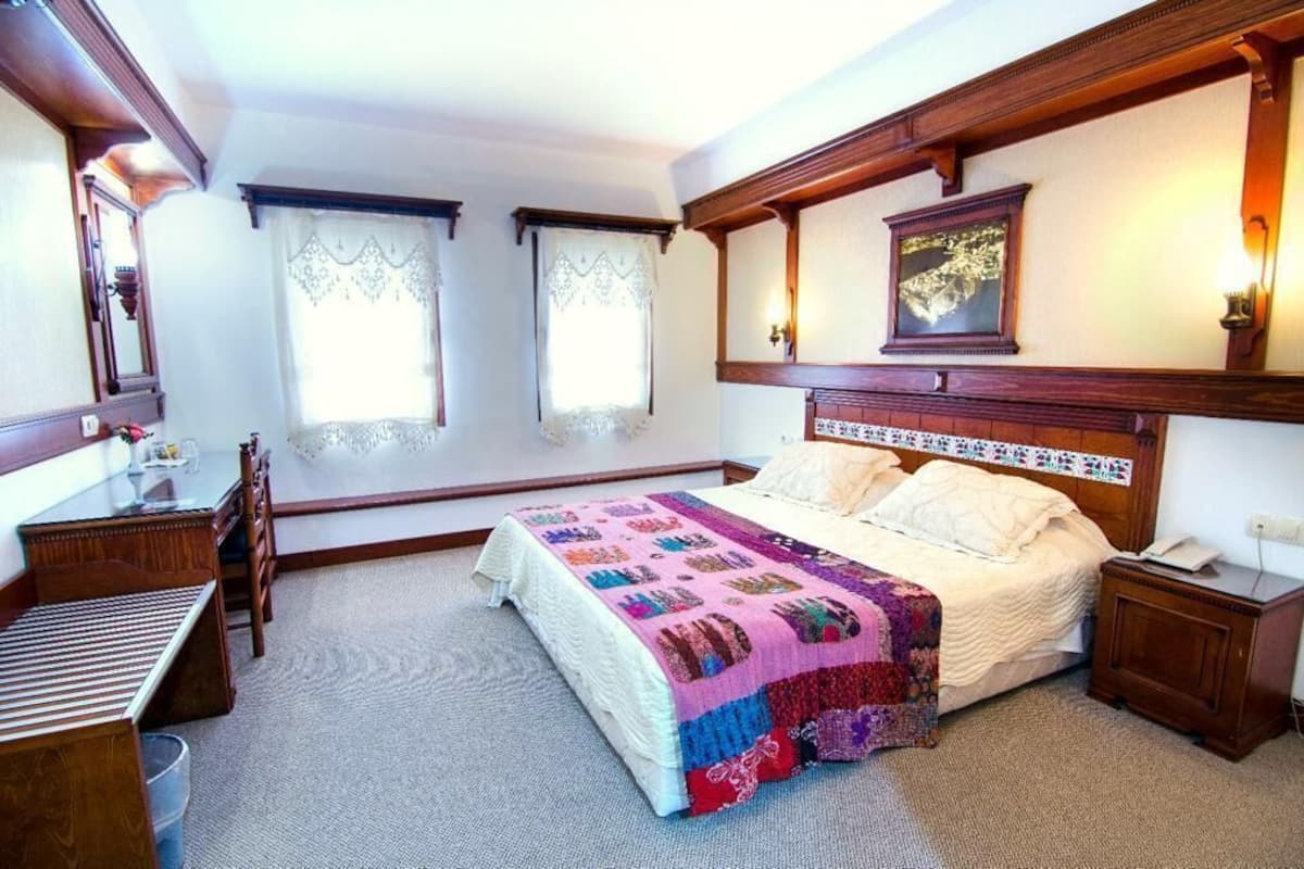 Quarto com cama, escrivaninha, duas janelas com cortinas, mesa de cabeceira e luminária na parede.