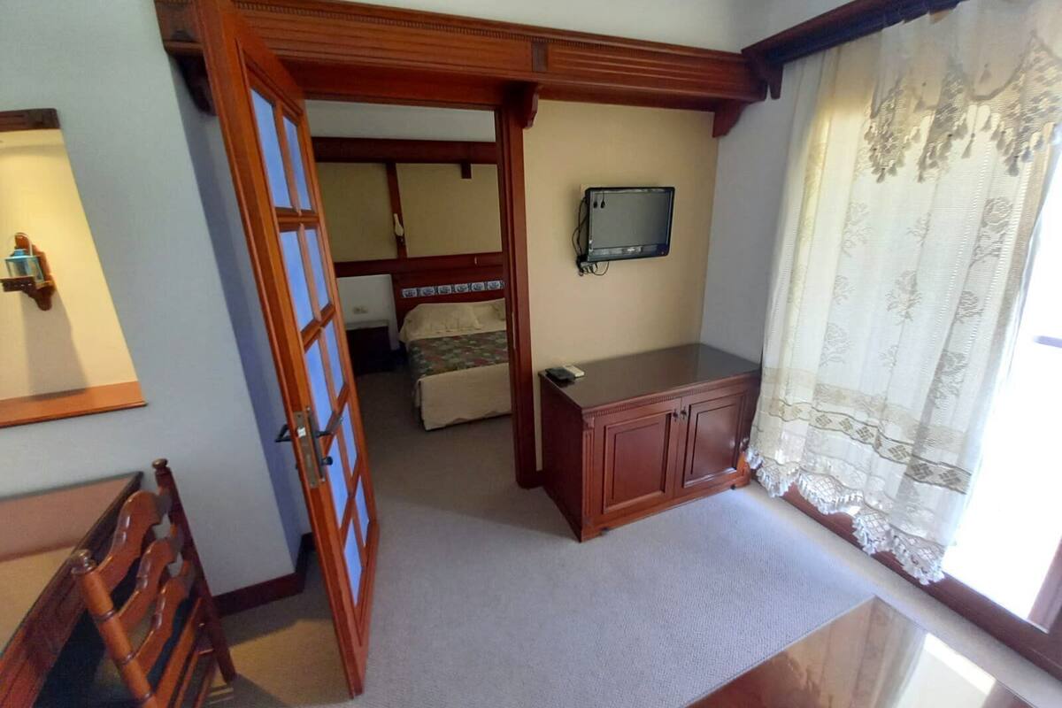 Quarto com guarda-roupa de madeira, televisão fixada na parede, cama com colcha floral e porta entreaberta para outro espaço.