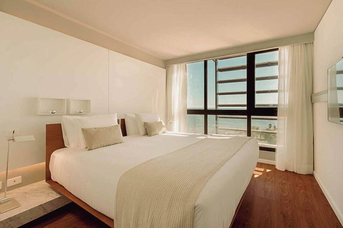 Quarto de hotel moderno com uma cama grande, uma mesa de cabeceira, um abajur e vista para o mar através de uma grande janela.