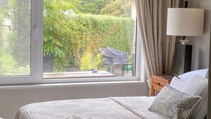 Quarto com uma janela grande, cama com roupa de cama estampada, mesa de cabeceira com abajur e vista para o verde lá fora.
