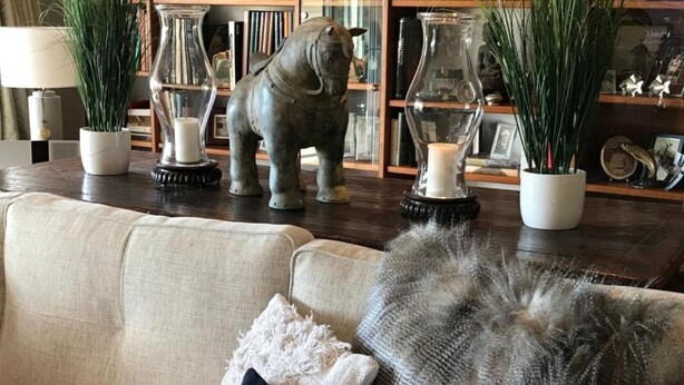 Sala de estar com um sofá bege, almofada azul escura e manta felpuda. Há duas plantas em vasos, um vaso de vidro e uma escultura de metal sobre a mesa de centro.