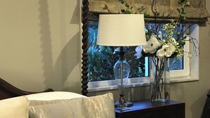 Um quarto com uma cama, uma mesa de cabeceira com abajur e um vaso de flores, e uma janela com vista para o verde.