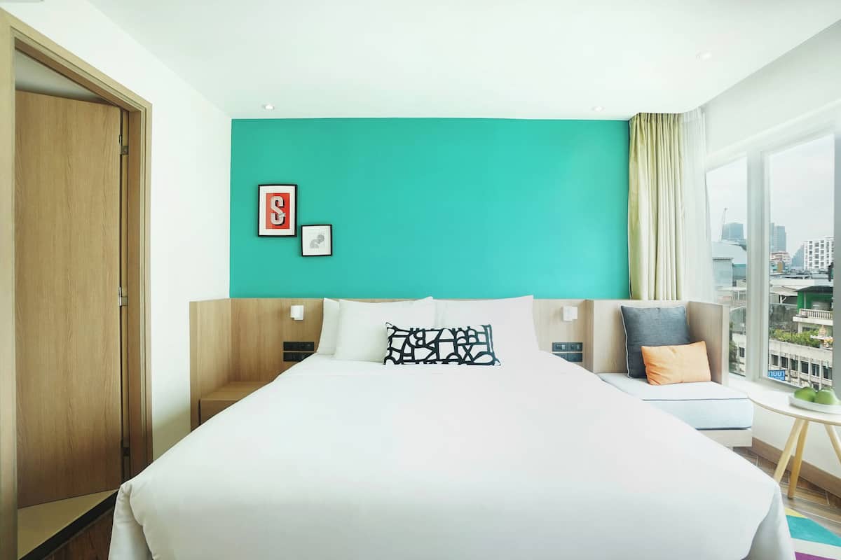 Quarto de hotel com uma cama grande, uma parede de destaque azul-esverdeada, vista para a cidade, um guarda-roupa de madeira e uma pequena mesa com uma planta.