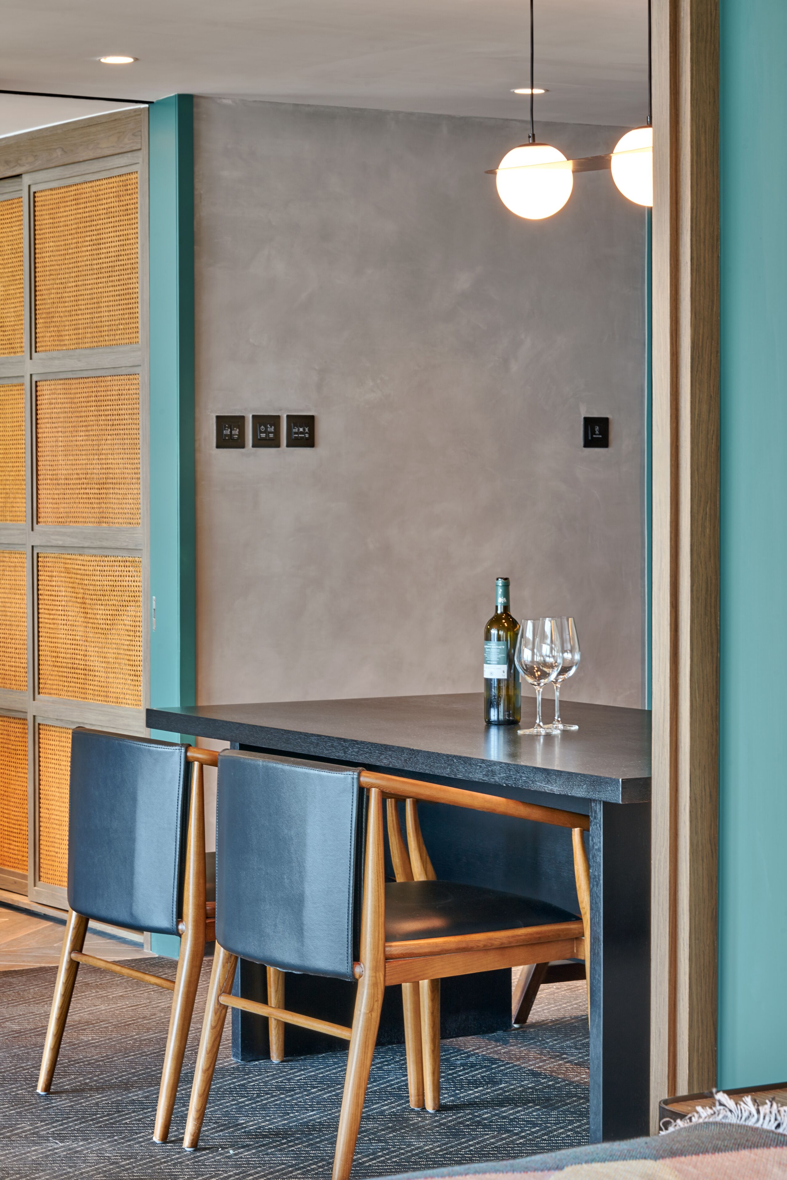 Uma área de bar moderna com bancada escura, duas cadeiras de madeira, uma garrafa de vinho e dois copos.
