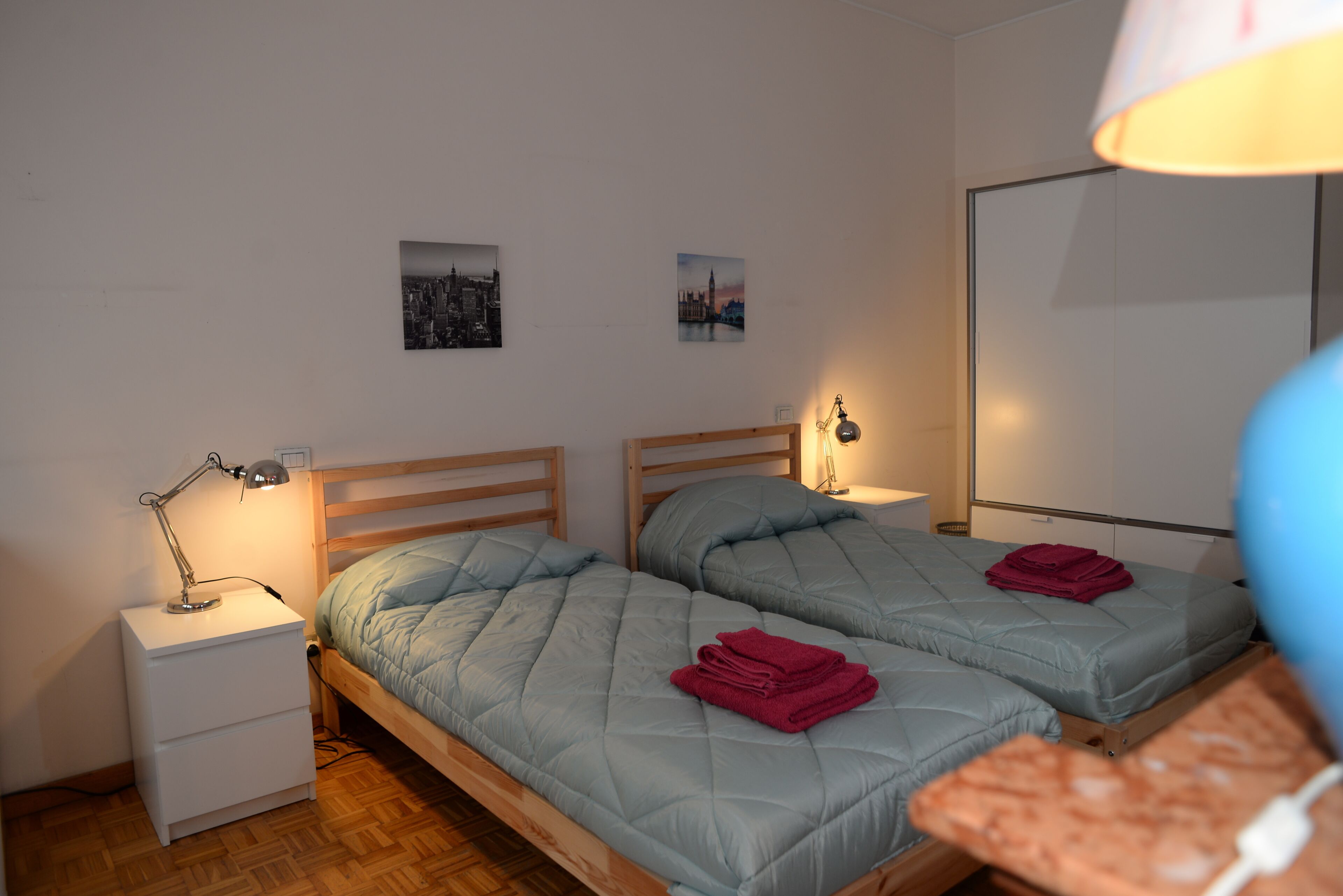 Quarto com duas camas, uma mesa de cabeceira com luminária e duas imagens penduradas na parede.