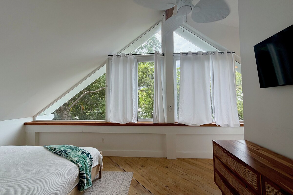 Quarto com teto inclinado, cama com manta verde e branca estampada, cômoda de madeira e janela com cortinas brancas.