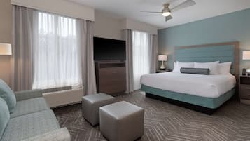 Quarto de hotel com cama, televisão, ventoinha no teto e uma grande janela com cortinas.