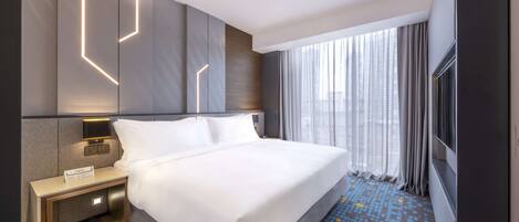 Quarto de hotel moderno com uma cama grande, uma mesinha de cabeceira com abajur, televisão e janela com cortinas.
