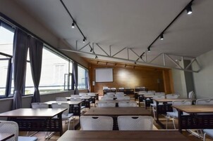 Sala de conferências com mesas e cadeiras dispostas para reuniões, uma tela de projetor e grandes janelas.