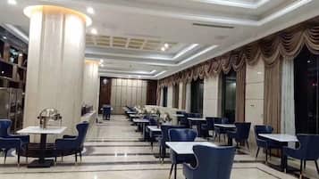 Área de refeições de hotel com mesas e cadeiras dispostas em fileiras, piso de mármore e um corredor longo que leva à saída.
