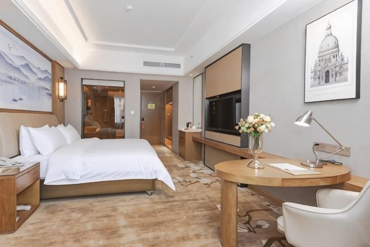 Quarto de hotel com cama, escrivaninha, cadeira, televisão e obras de arte nas paredes.