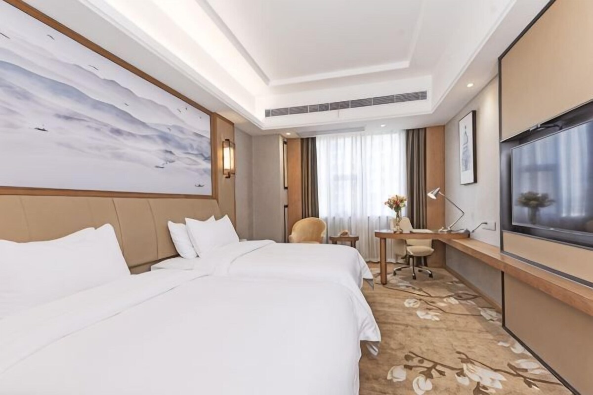 Quarto de hotel com duas camas, uma escrivaninha, uma cadeira, uma televisão e um quadro grande na parede.