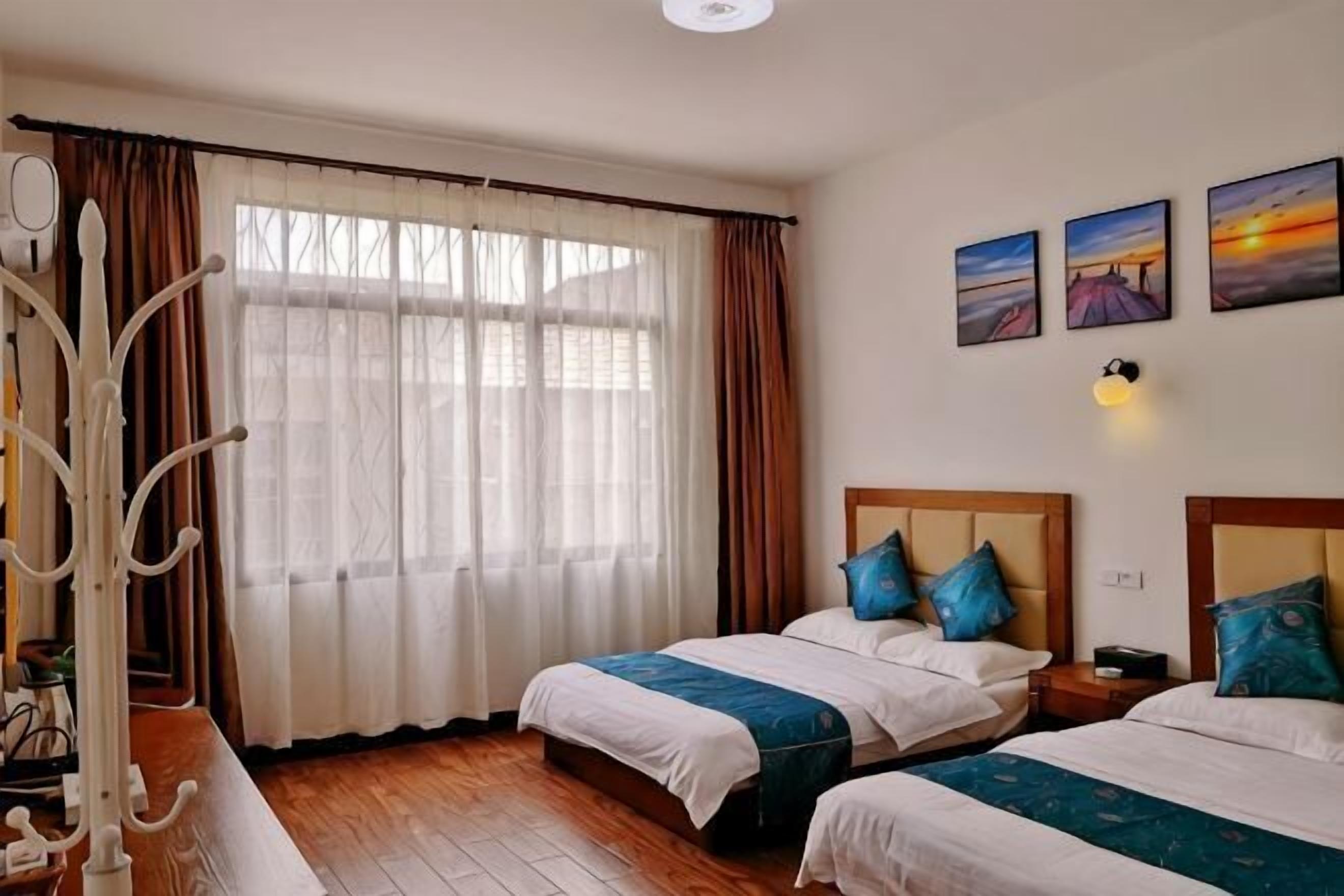 Quarto de hotel com duas camas, uma janela grande com cortinas, um porta-roupas e obras de arte nas paredes.