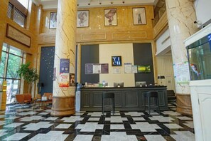 Um hall de entrada com balcão de recepção, balcões de check-in e check-out. Há cadeiras e um vaso de planta.