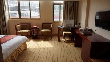 Quarto de hotel com cama, duas cadeiras, escrivaninha com computador, televisão e amplas janelas com cortinas.