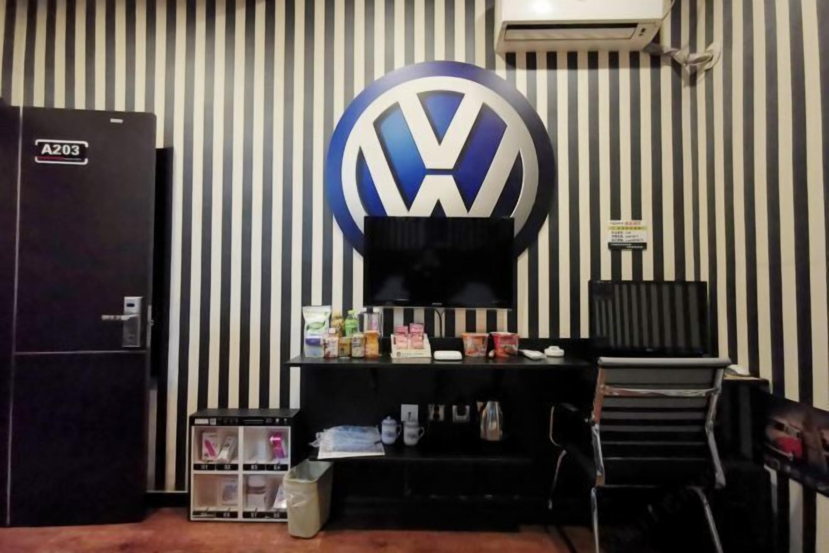 Um quarto com parede listrada, uma geladeira preta modelo A203, uma escrivaninha com diversos objetos, e o logotipo da VW na parede.