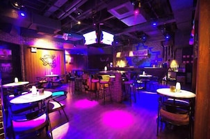 Um bar com várias mesas e banquetas, parede de tijolos com letreiro de neon e palco com equipamento de DJ.