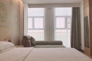 Quarto de hotel com uma cama grande, área de estar, mesa de cabeceira e janela com cortinas.