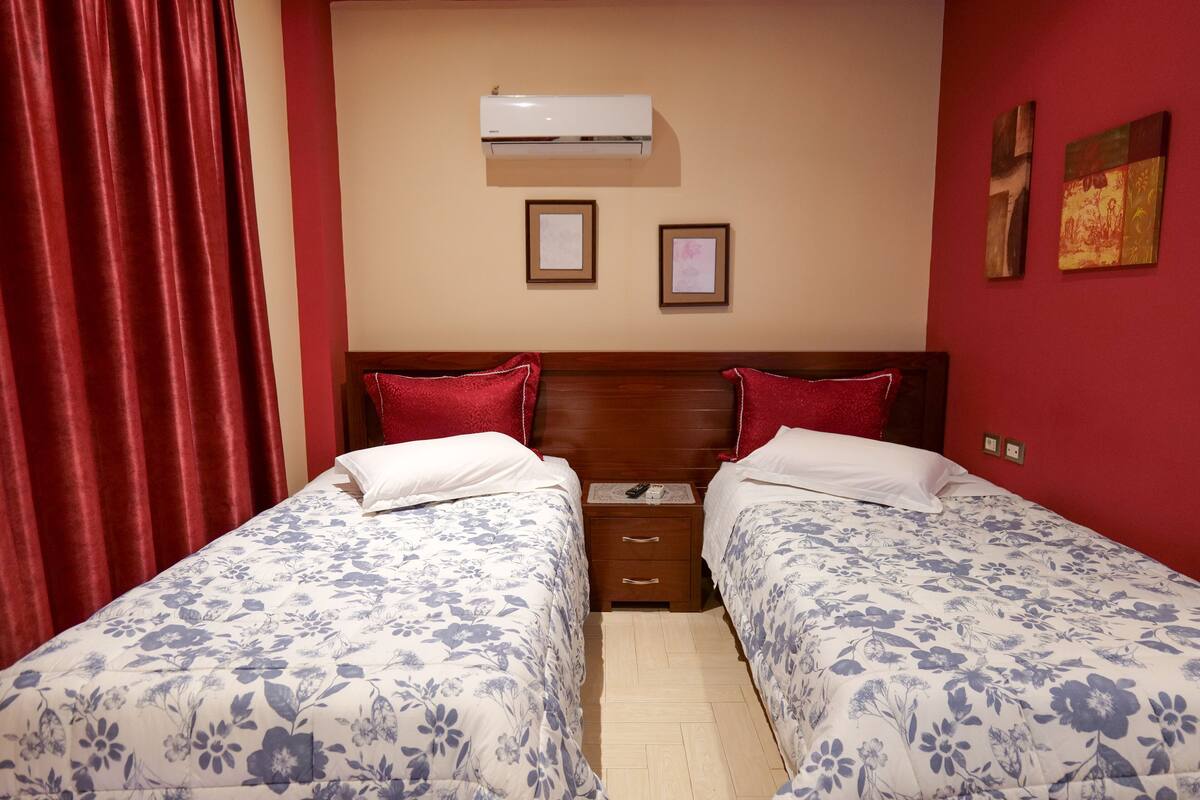 Quarto de hotel com duas camas, uma cabeceira de madeira, cortina vermelha e ar condicionado fixado na parede.