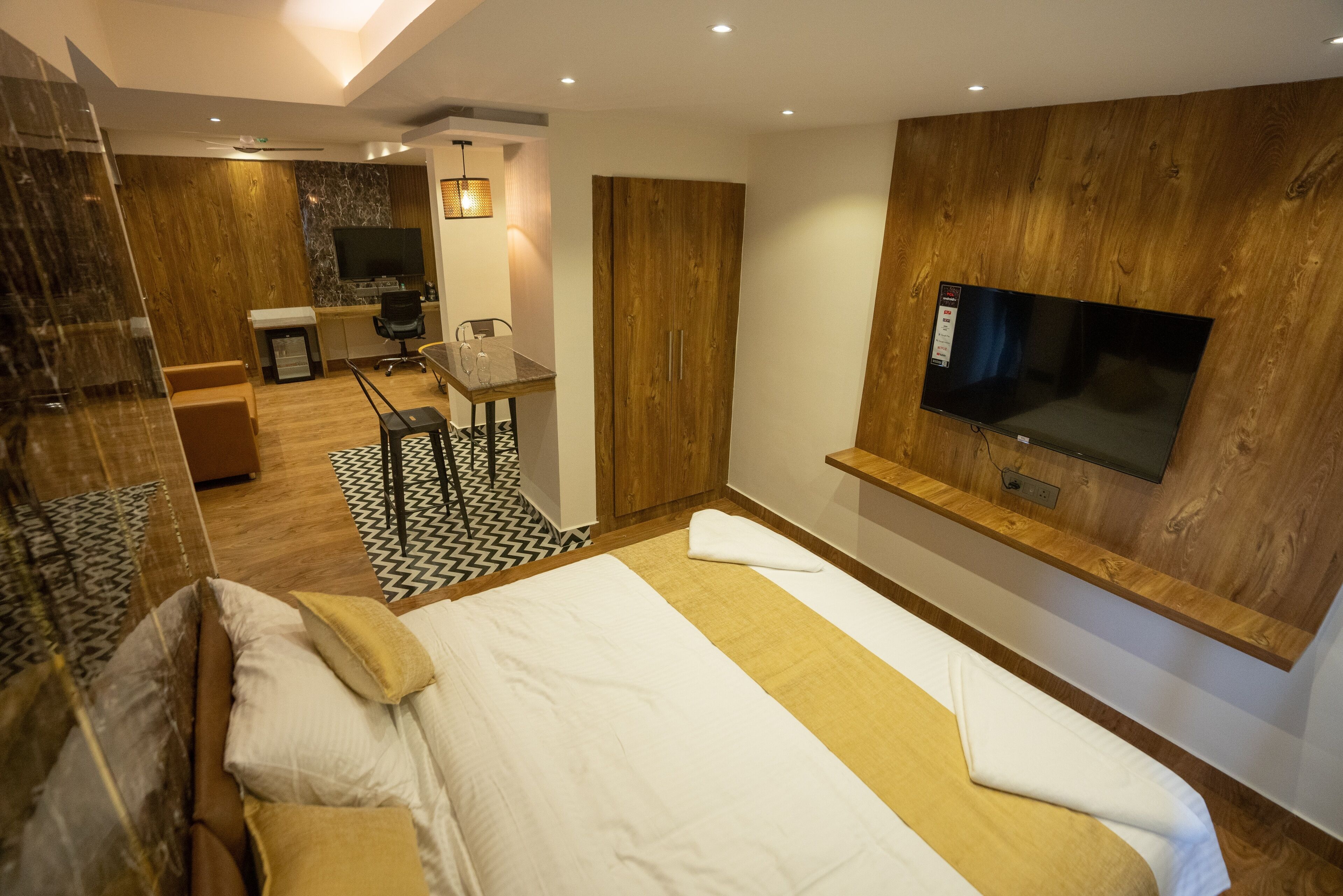 Quarto de hotel moderno com uma cama grande, uma TV fixada na parede, uma mesa de trabalho com cadeira e um guarda-roupa de madeira.