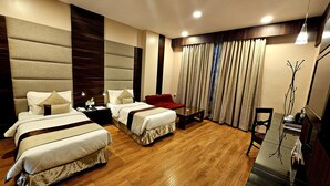 Quarto de hotel com duas camas, uma escrivaninha, uma cadeira e uma janela grande com cortinas.