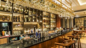 Uma área de bar com um balcão comprido, várias prateleiras com garrafas de bebidas, copos pendurados e alguns bancos.