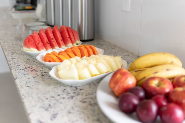 Uma bancada de cozinha com diversas frutas, incluindo melancia, laranjas, bananas, maçãs e ameixas.