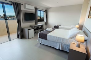 Quarto de hotel com cama, escrivaninha, cadeira, televisão e varanda com cortinas.