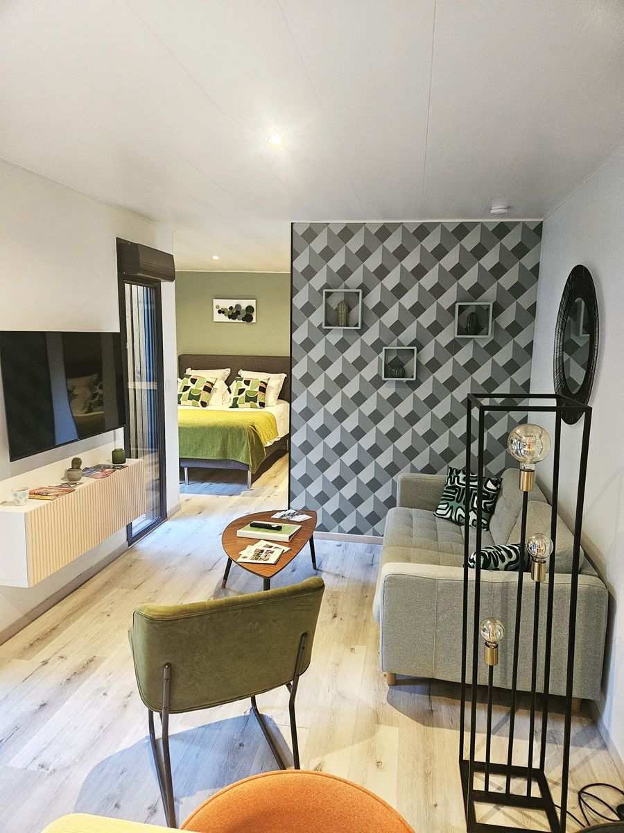 Uma sala de estar moderna com sofá, poltrona e mesa de centro. Há uma cama com colcha verde ao fundo.